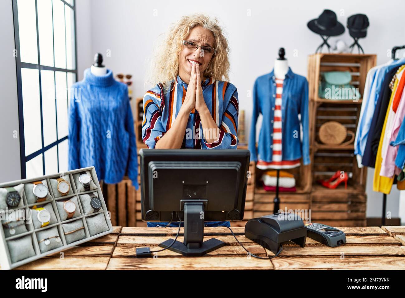 Middle age blonde woman working as manager at retail boutique praying with hands together asking ...