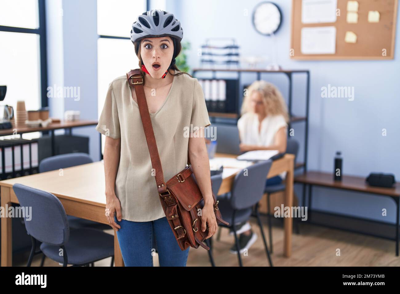 Young brunette woman working at the office wearing bike helmet scared ...
