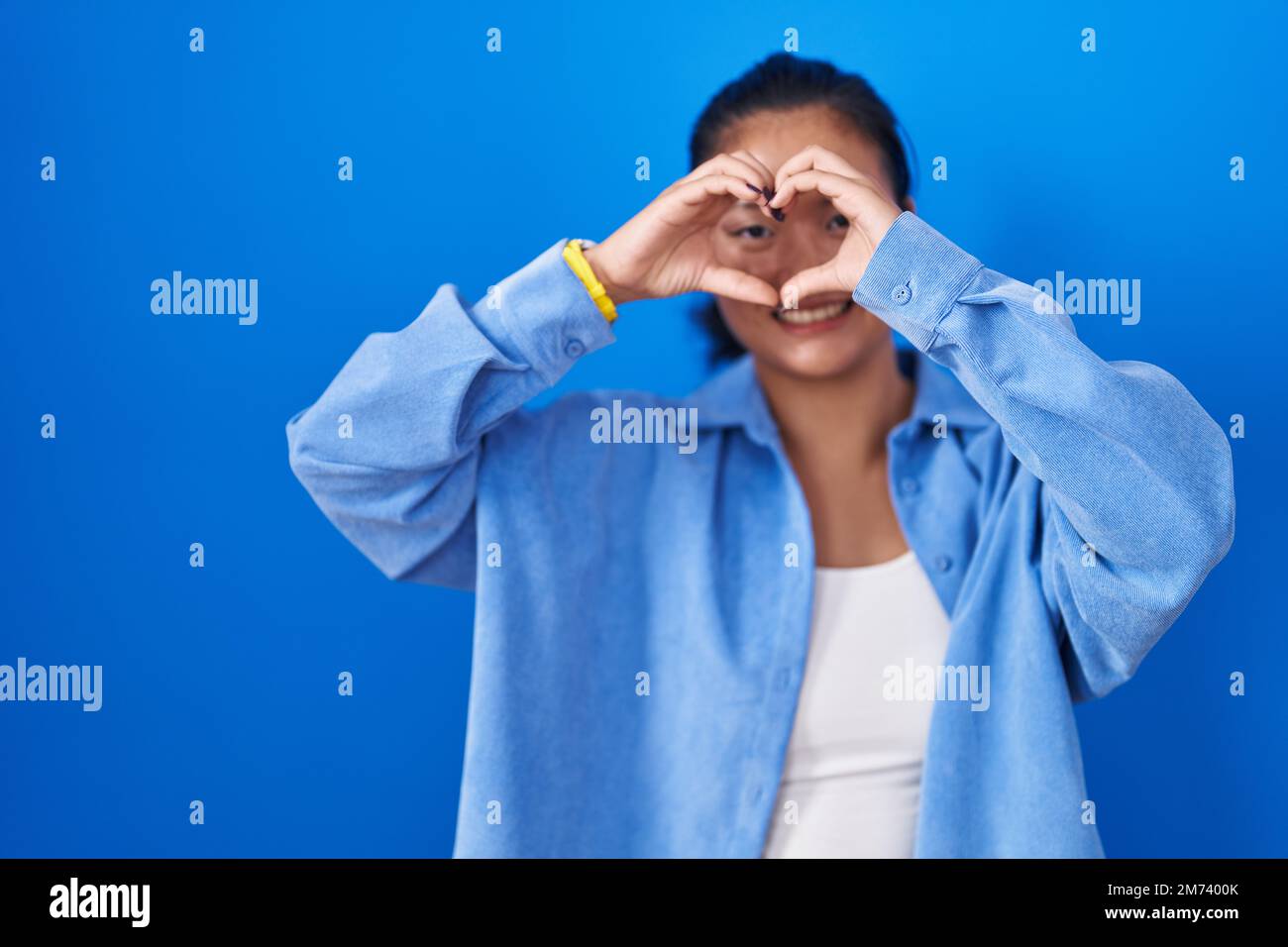 Asian young woman standing over blue background doing heart shape with ...