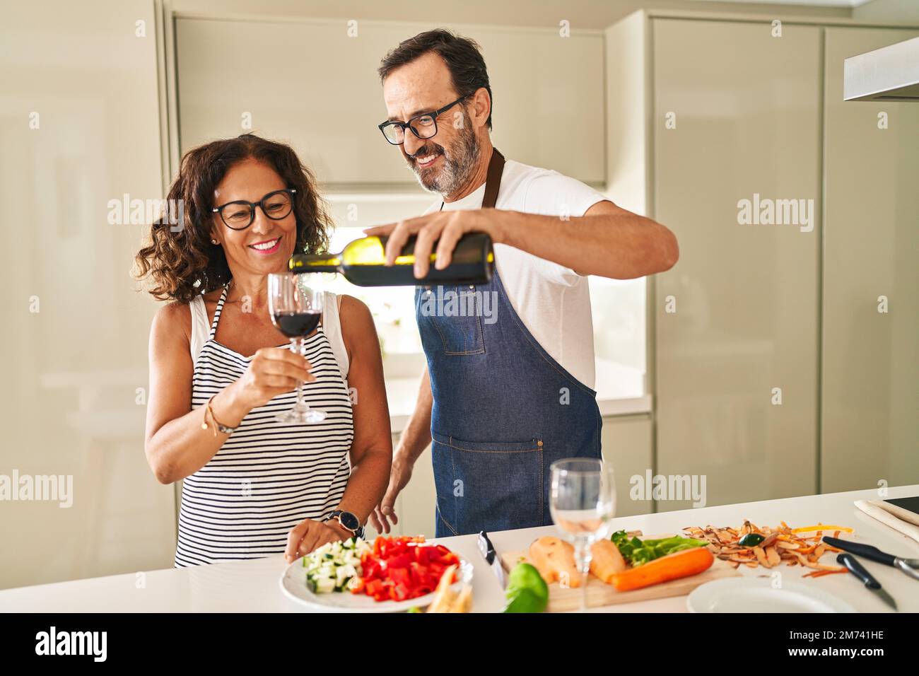 Middle age hispanic couple cooking and pouring wine on glass at kitchen ...