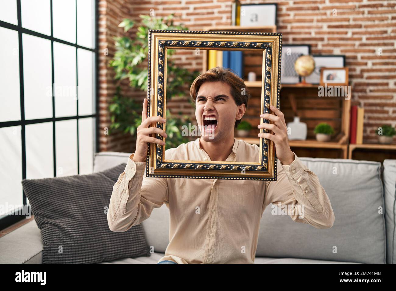 Young man holding empty frame angry and mad screaming frustrated and ...