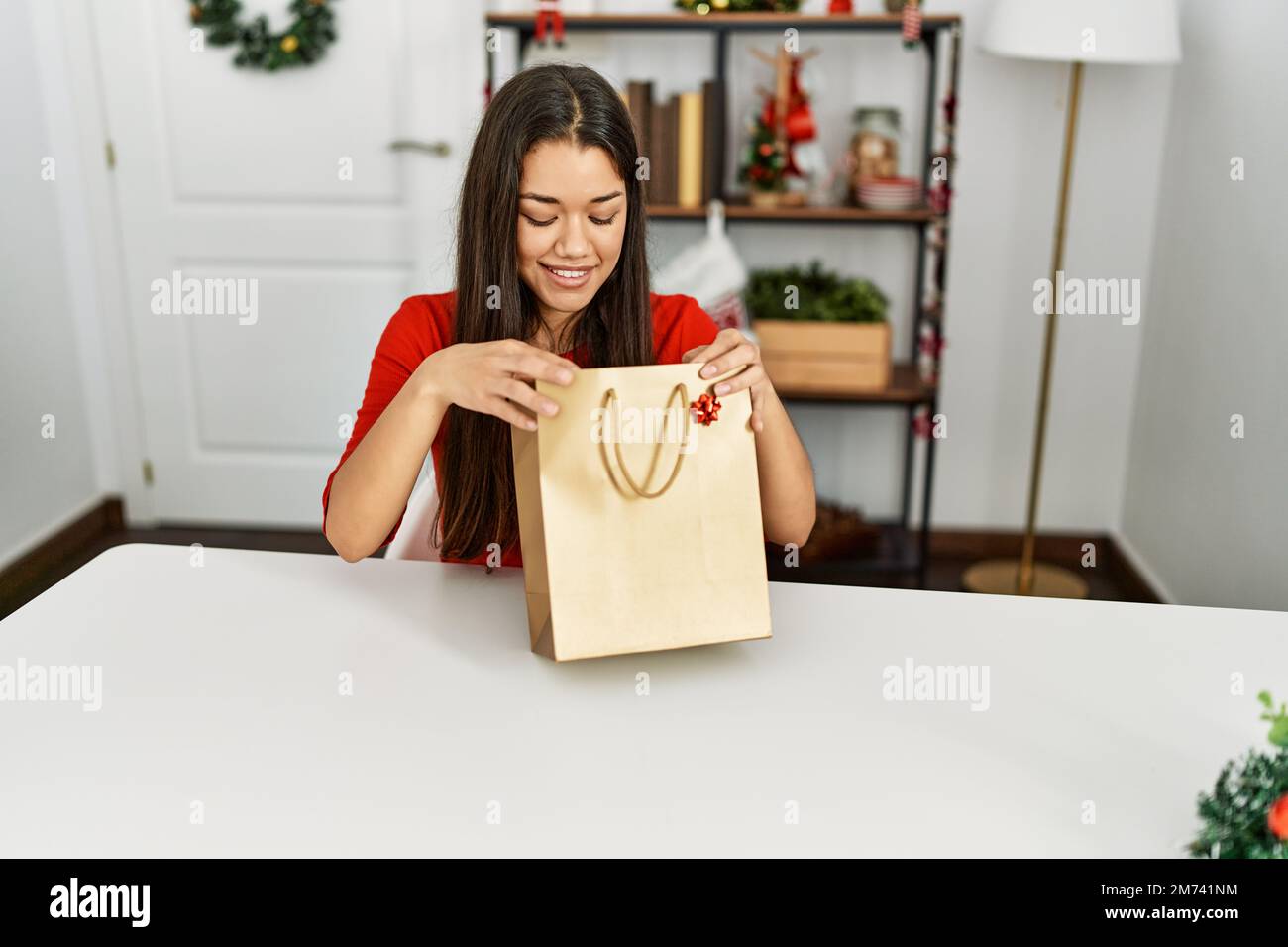 Young latin woman open gift bag sitting by christmas decor at home ...