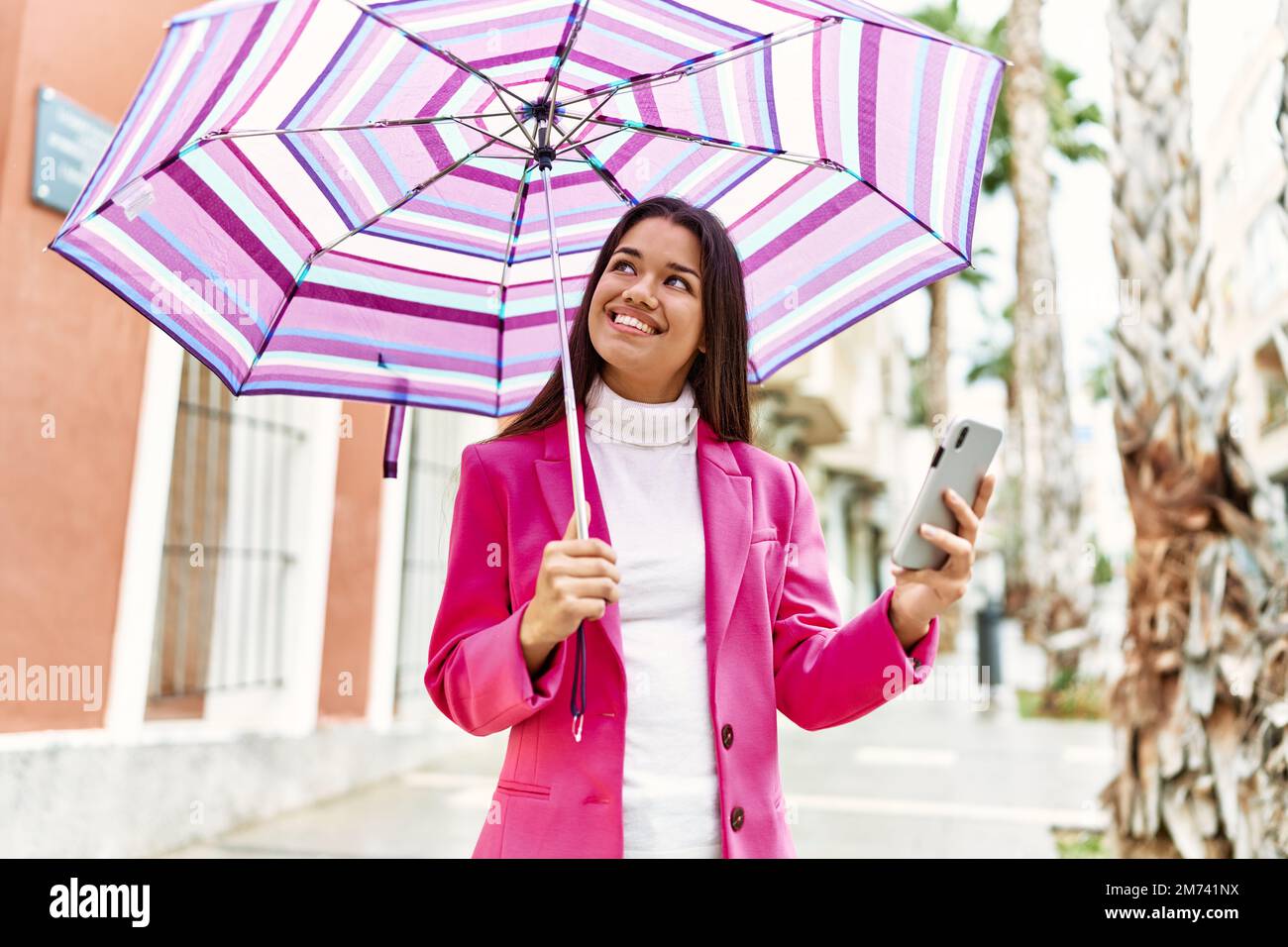 Young latin woman using smartphone holding umbrella at street Stock ...