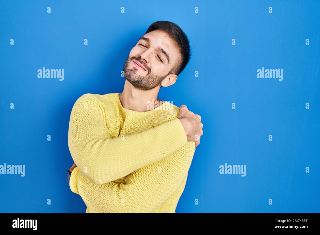 Hispanic man standing over blue background hugging oneself happy and ...
