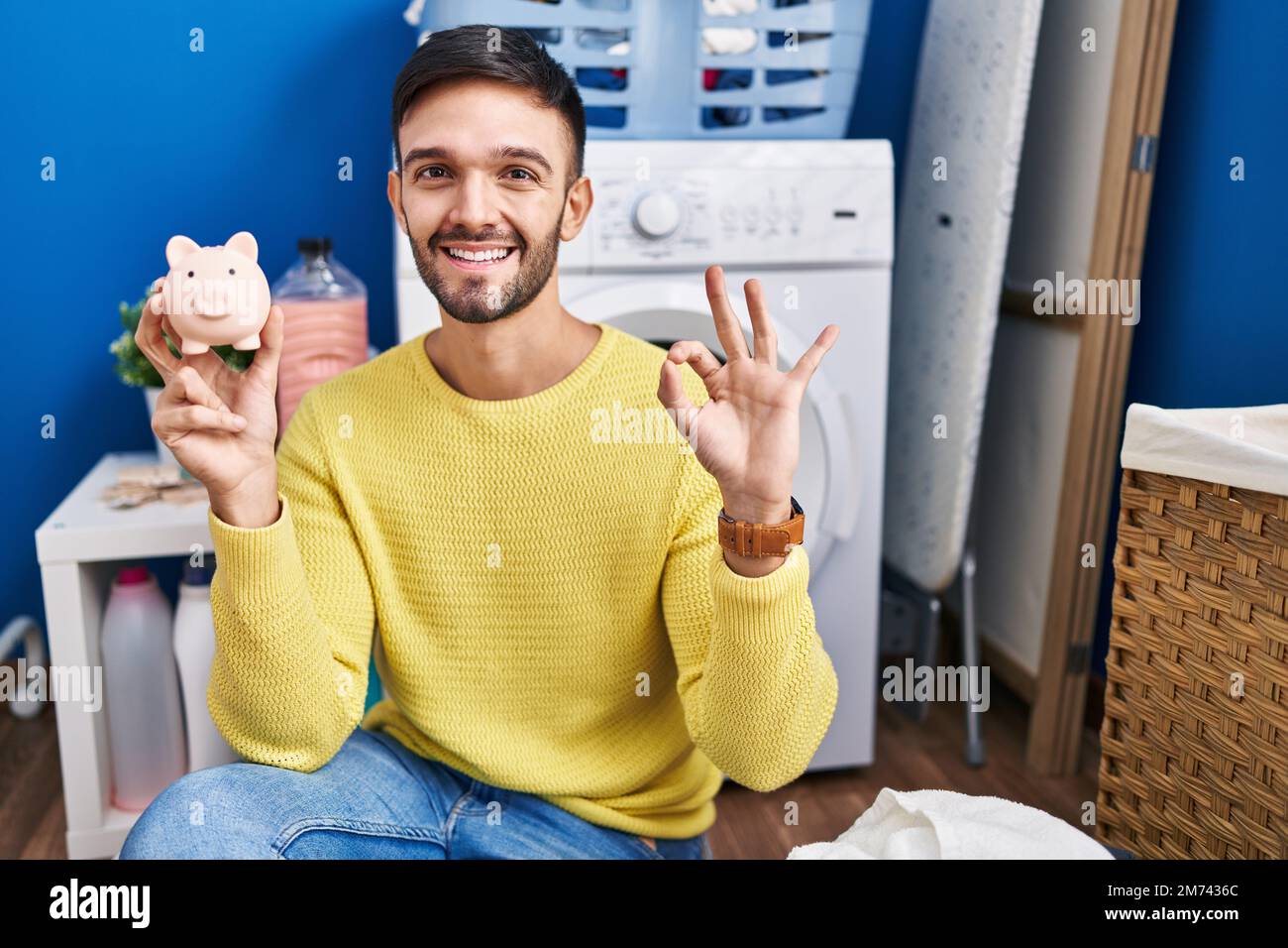 Hispanic man doing laundry holding piggy bank doing ok sign with ...