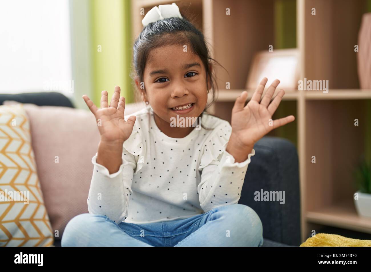 Adorable hispanic girl saying hello with hand sitting on sofa at home ...