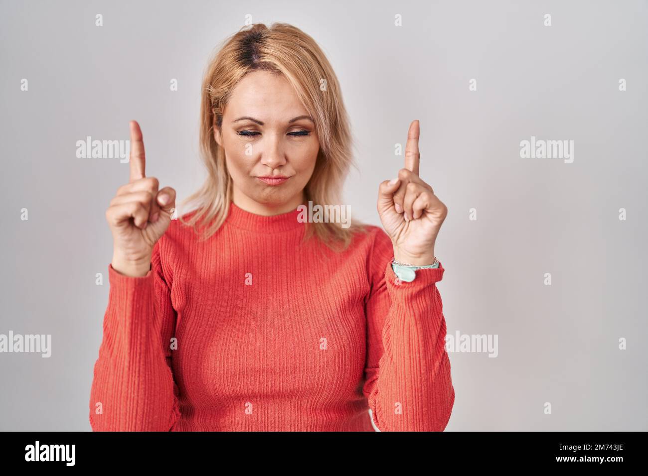 Blonde woman standing over isolated background pointing up looking sad ...