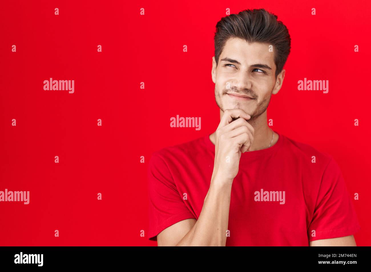 Young hispanic man standing over red background with hand on chin ...