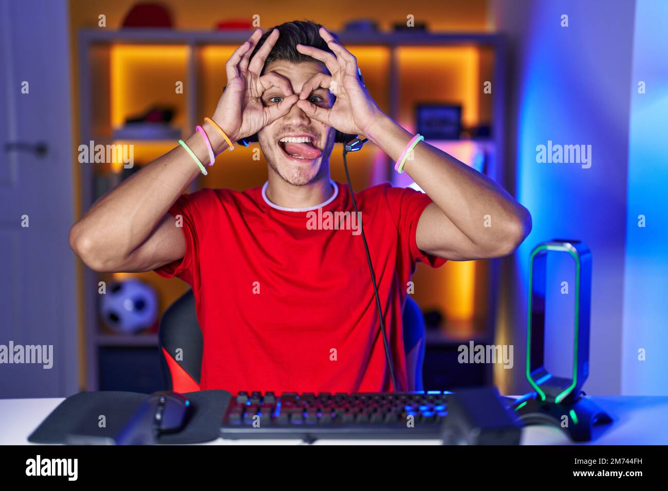 Young hispanic man playing video games doing ok gesture like binoculars ...