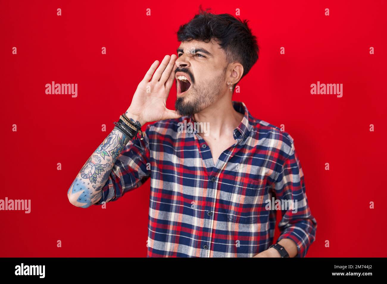 Young hispanic man with beard standing over red background shouting and ...