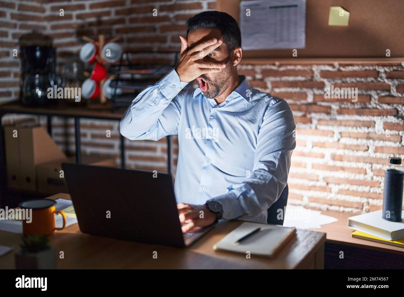 Hispanic man with beard working at the office at night peeking in shock ...