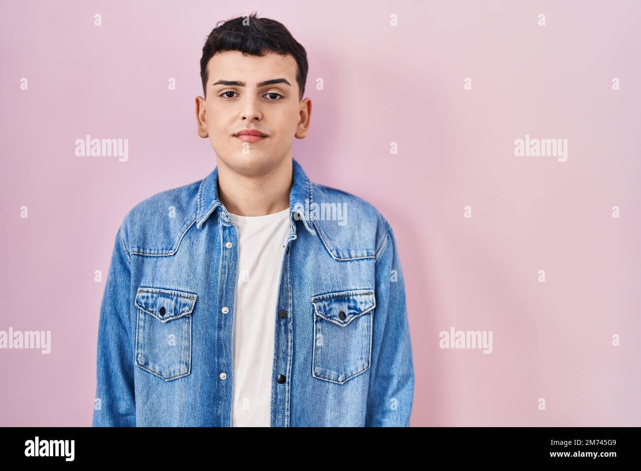 Non binary person standing over pink background with serious expression ...