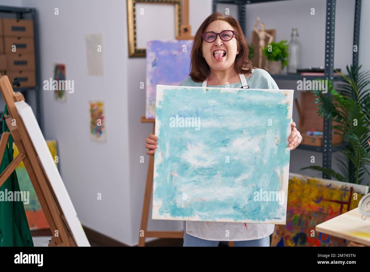 Senior woman with glasses holding canvas at art studio sticking tongue ...
