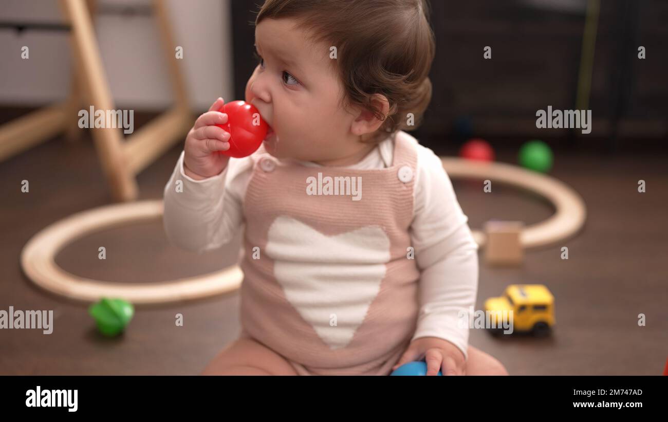 Adorable toddler sucking plastic ball sitting on floor at kindergarten ...