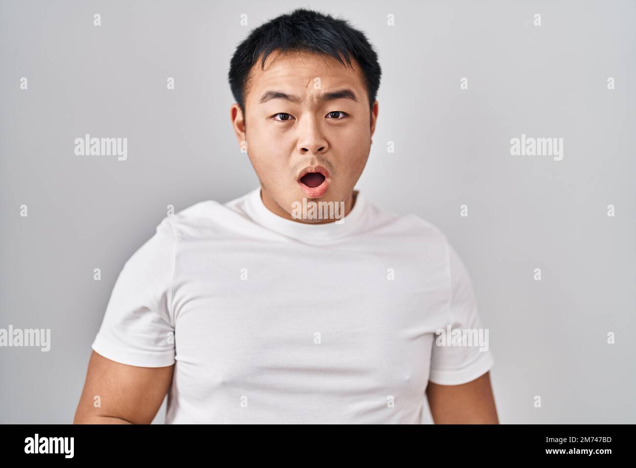 Young chinese man standing over white background in shock face, looking ...