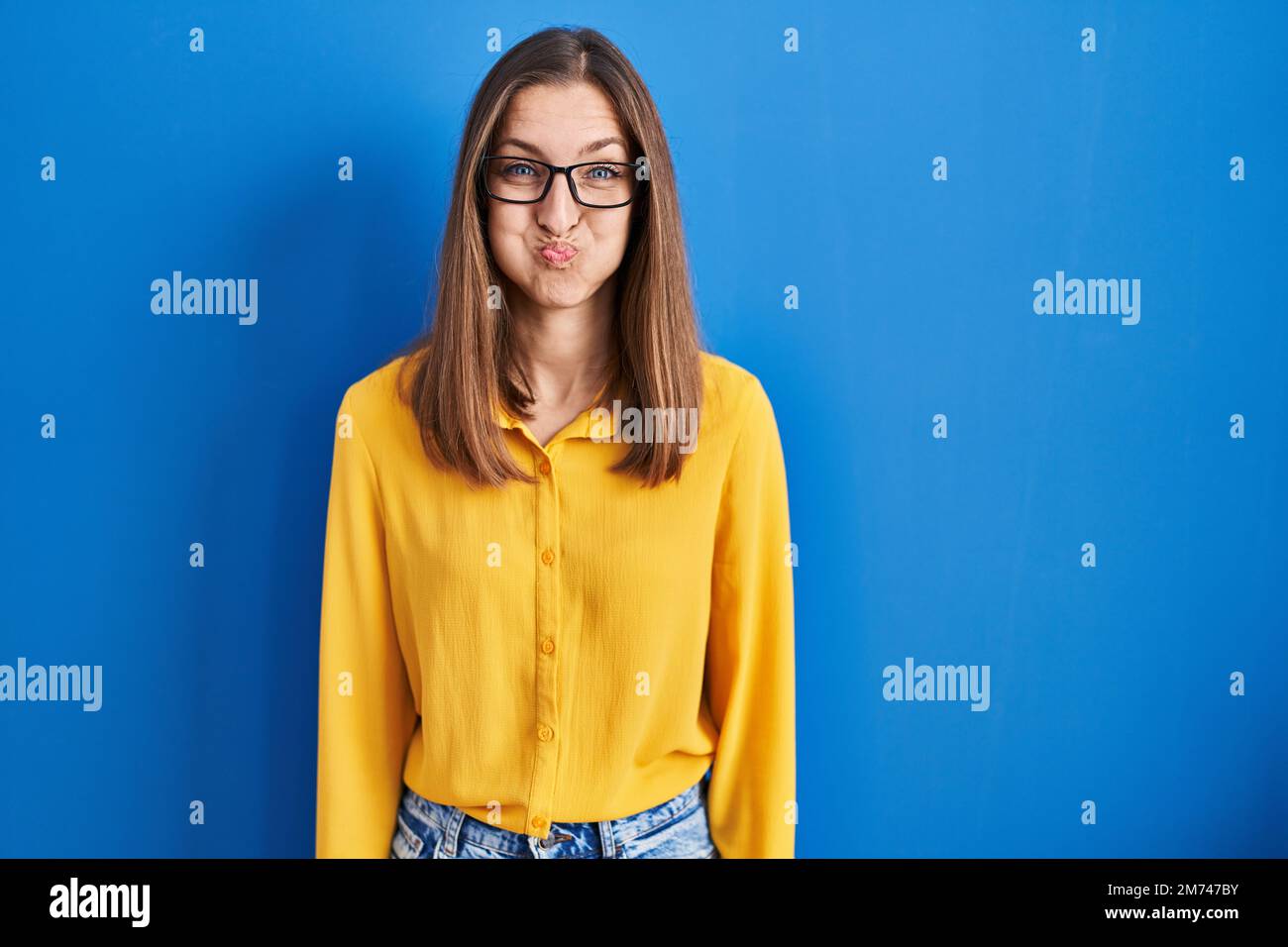 Young woman wearing glasses standing over blue background puffing ...