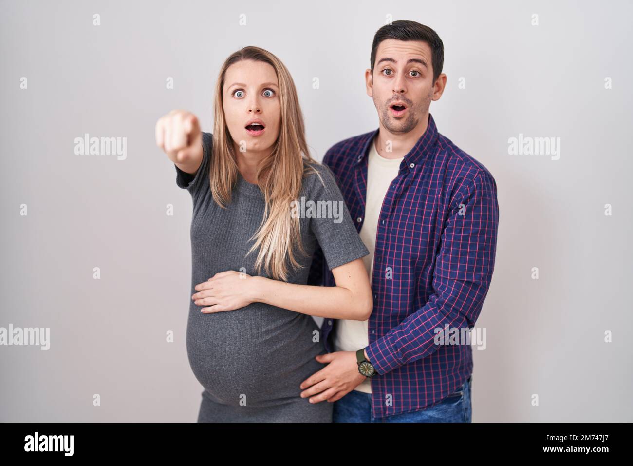 Young couple expecting a baby standing over white background pointing ...