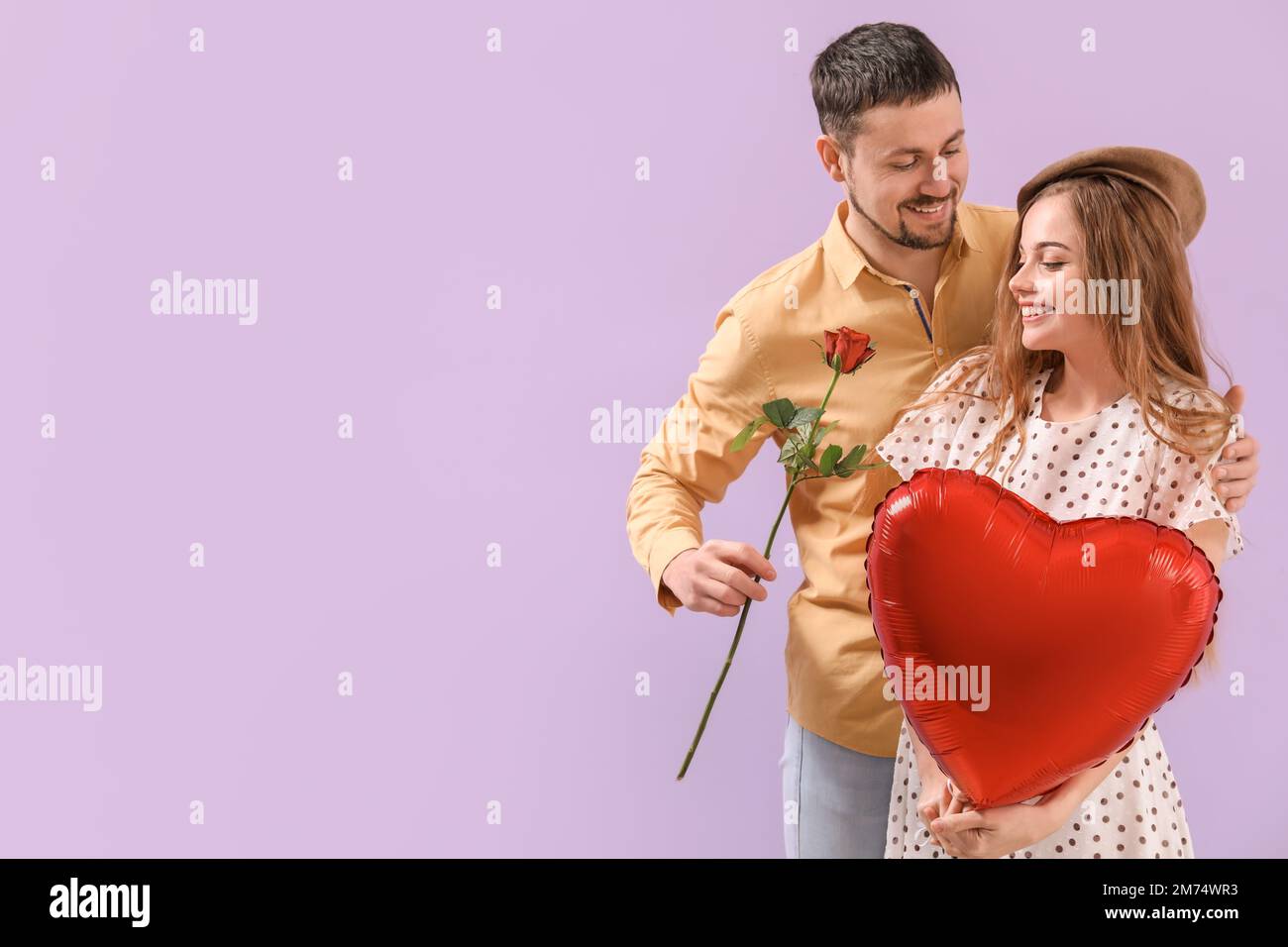 Happy young couple with heart-shaped balloon and flower on lilac ...