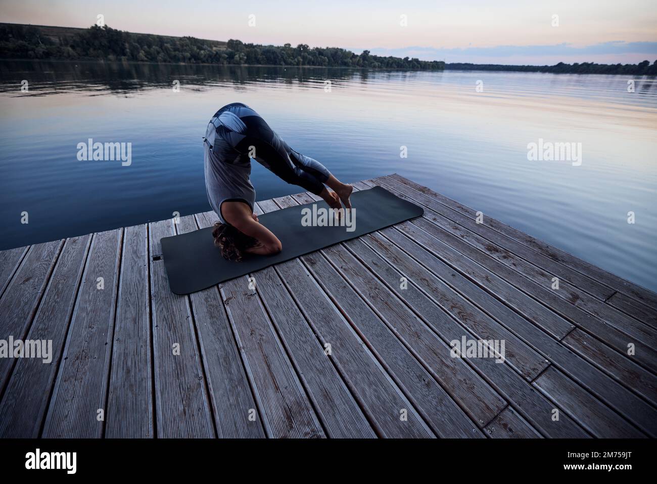 A fit woman practices yoga on the dock near the river and balances on ...