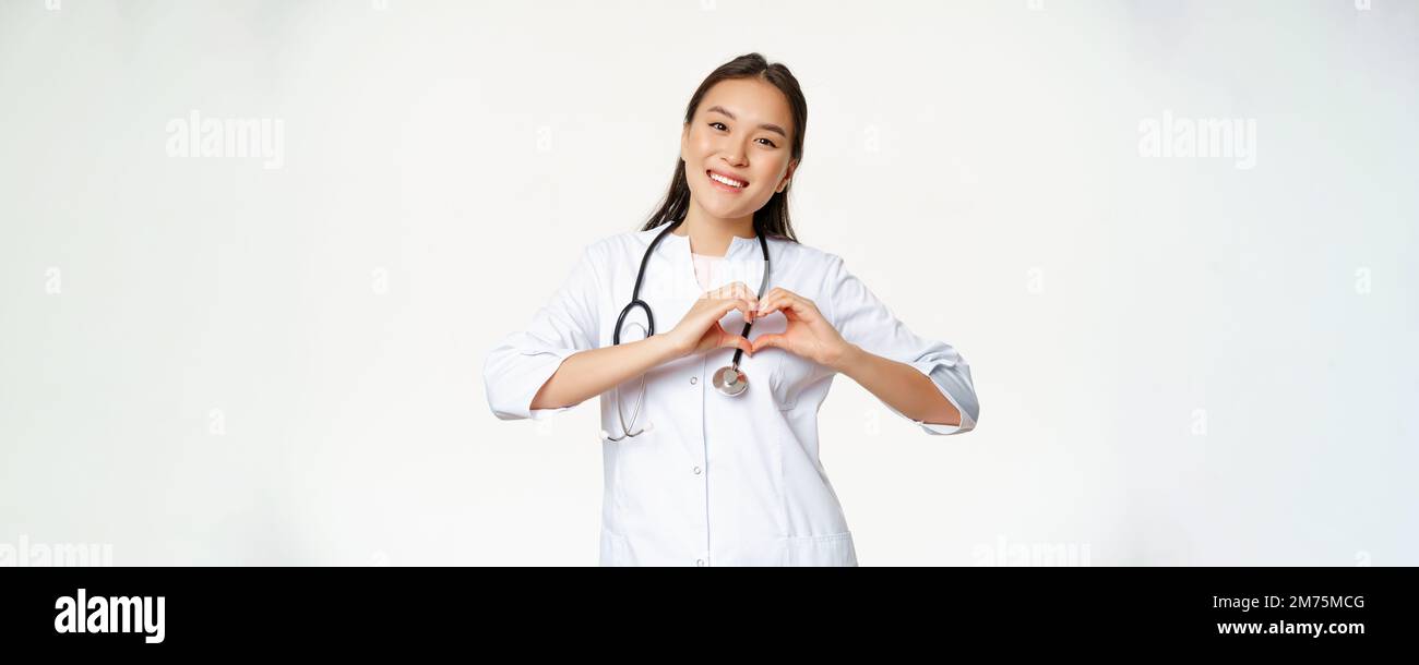 Smiling asian female doctor care for her patients, shows heart gesture ...