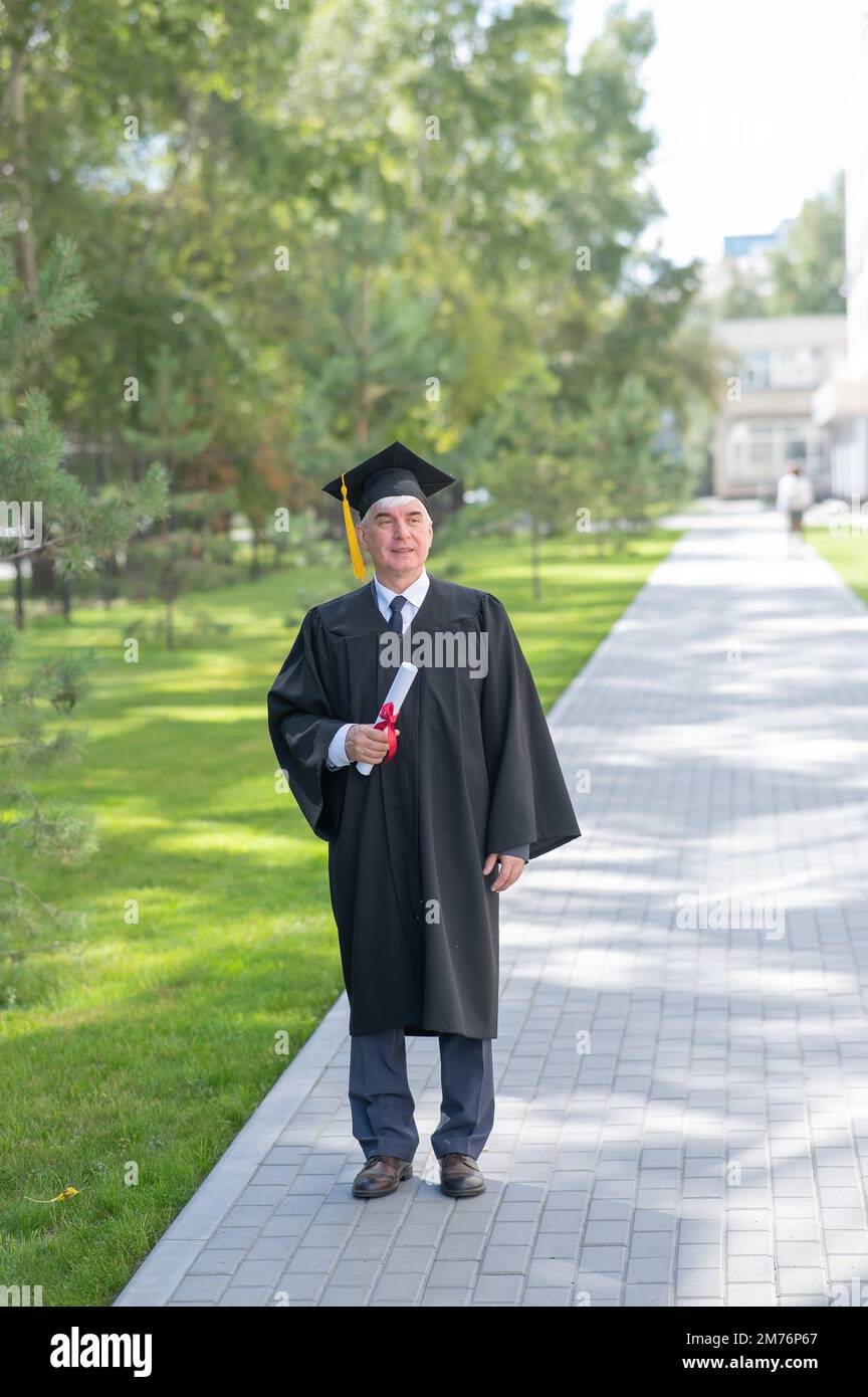 An old man in a graduation gown walks outdoors and holds a diploma ...