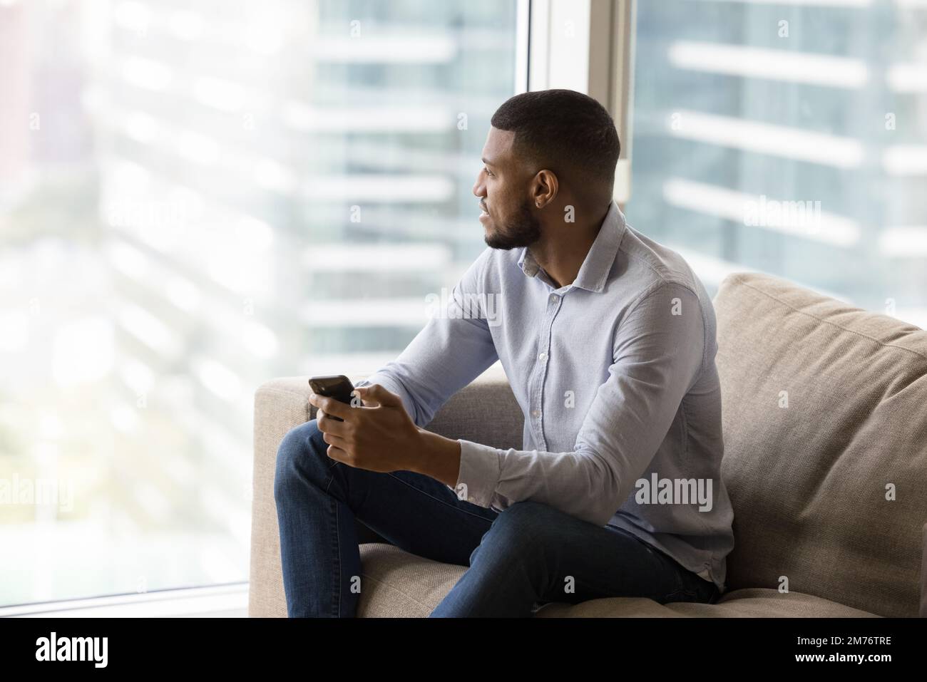 African man resting on couch holds smartphone staring out window Stock ...