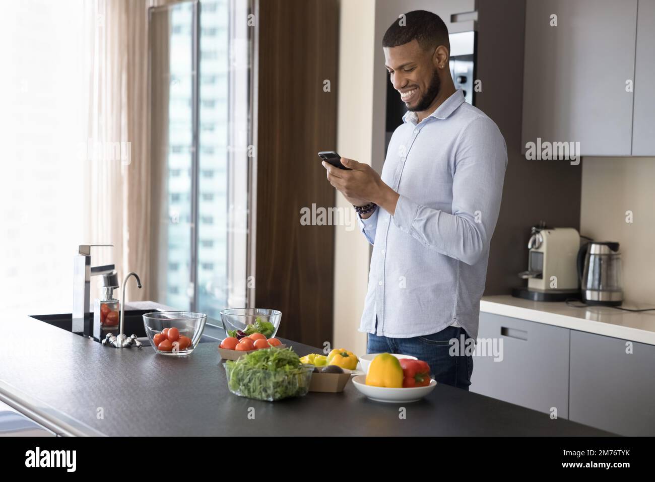 Young handsome African guy using cellphone standing in modern kitchen ...