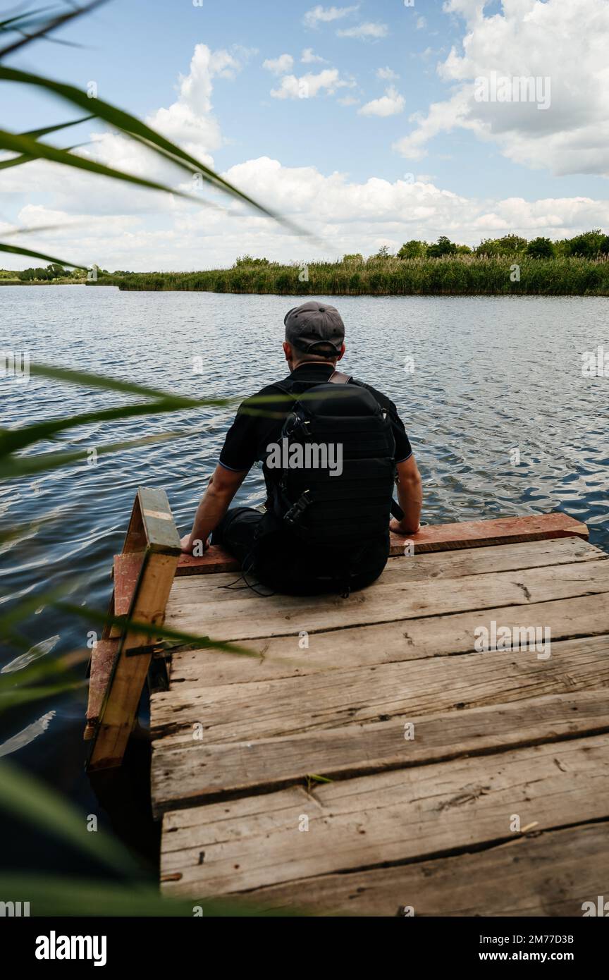 A man sits on the edge of a wooden bridge near a lake next to reeds, a ...