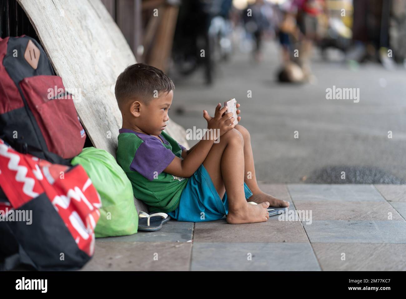 A young boy in the Philippines, sits on a pavement using a mobile phone ...