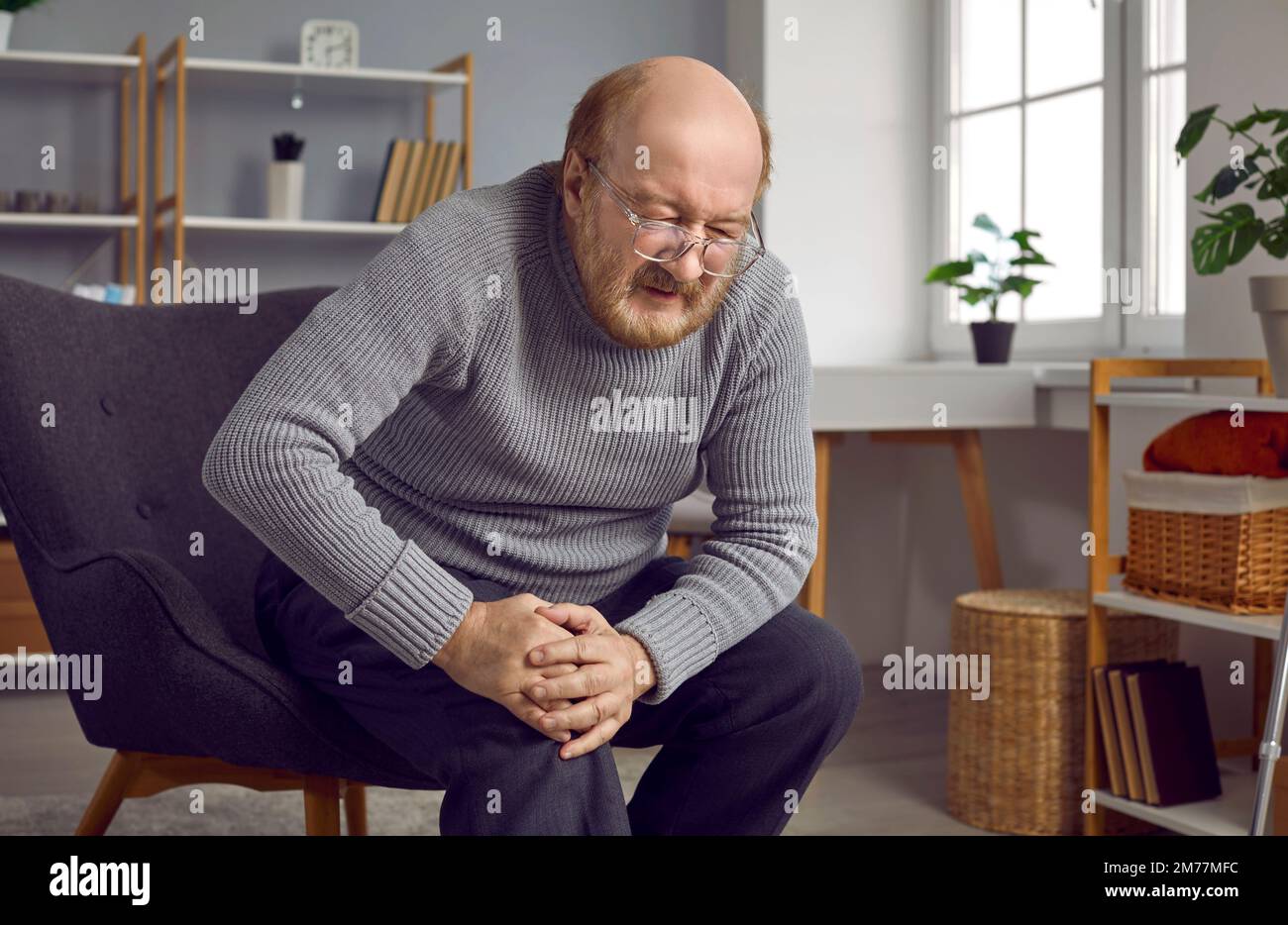 Senior man suffering from knee pain, sitting in his armchair and ...