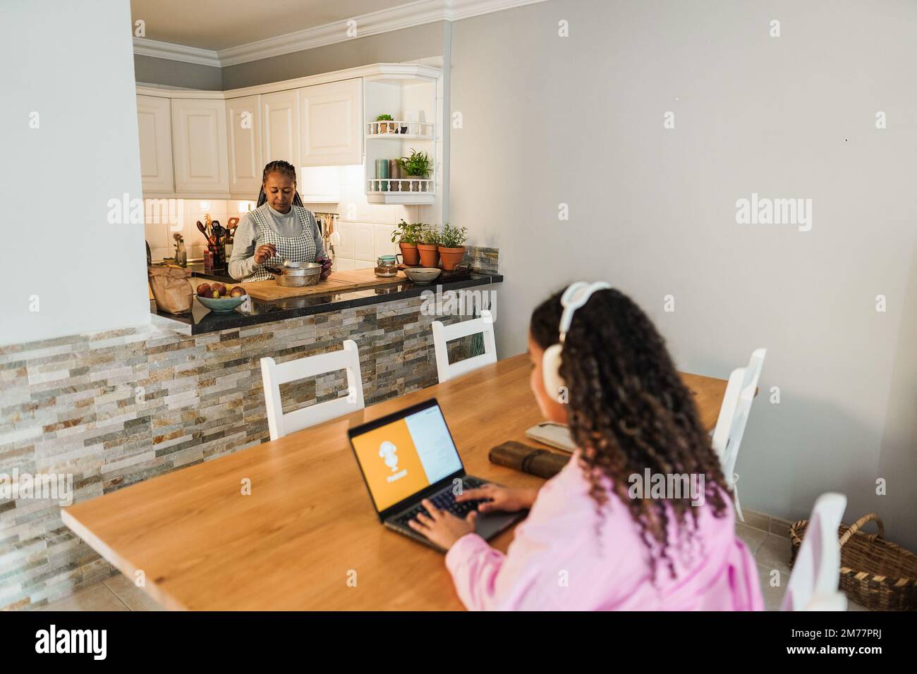 African daughter using laptop while mother cooking in kitchen at home ...