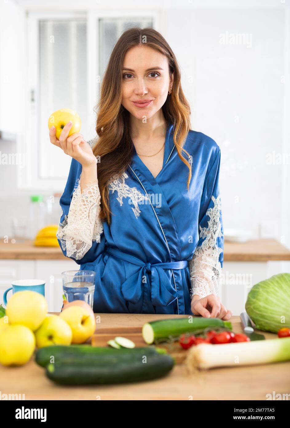 Satisfied woman in dressing gown eating apples in kitchen during ...