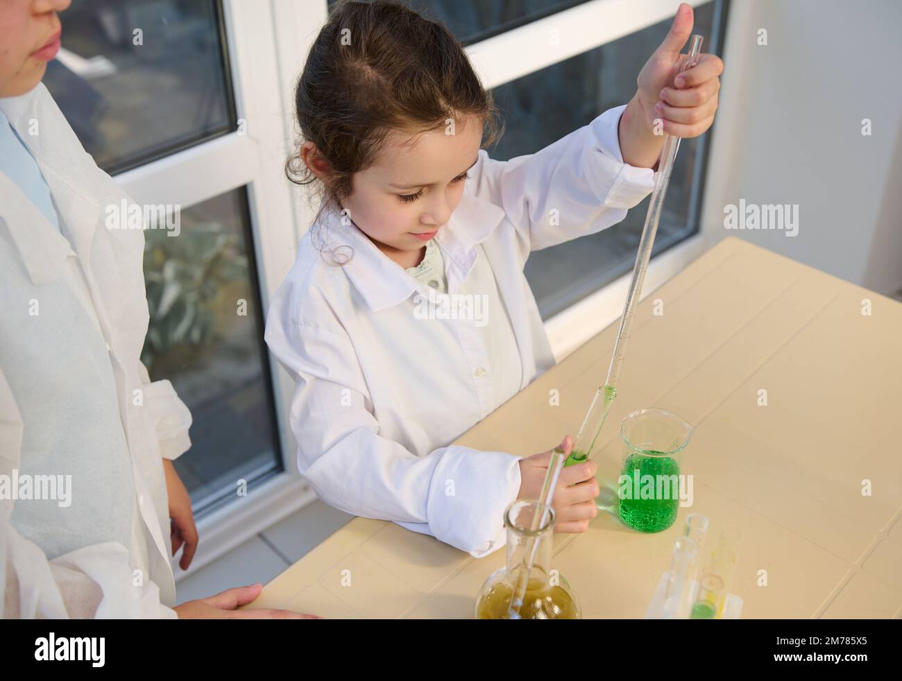 Smart little girl in lab coat, filling a graduated pipette with green ...