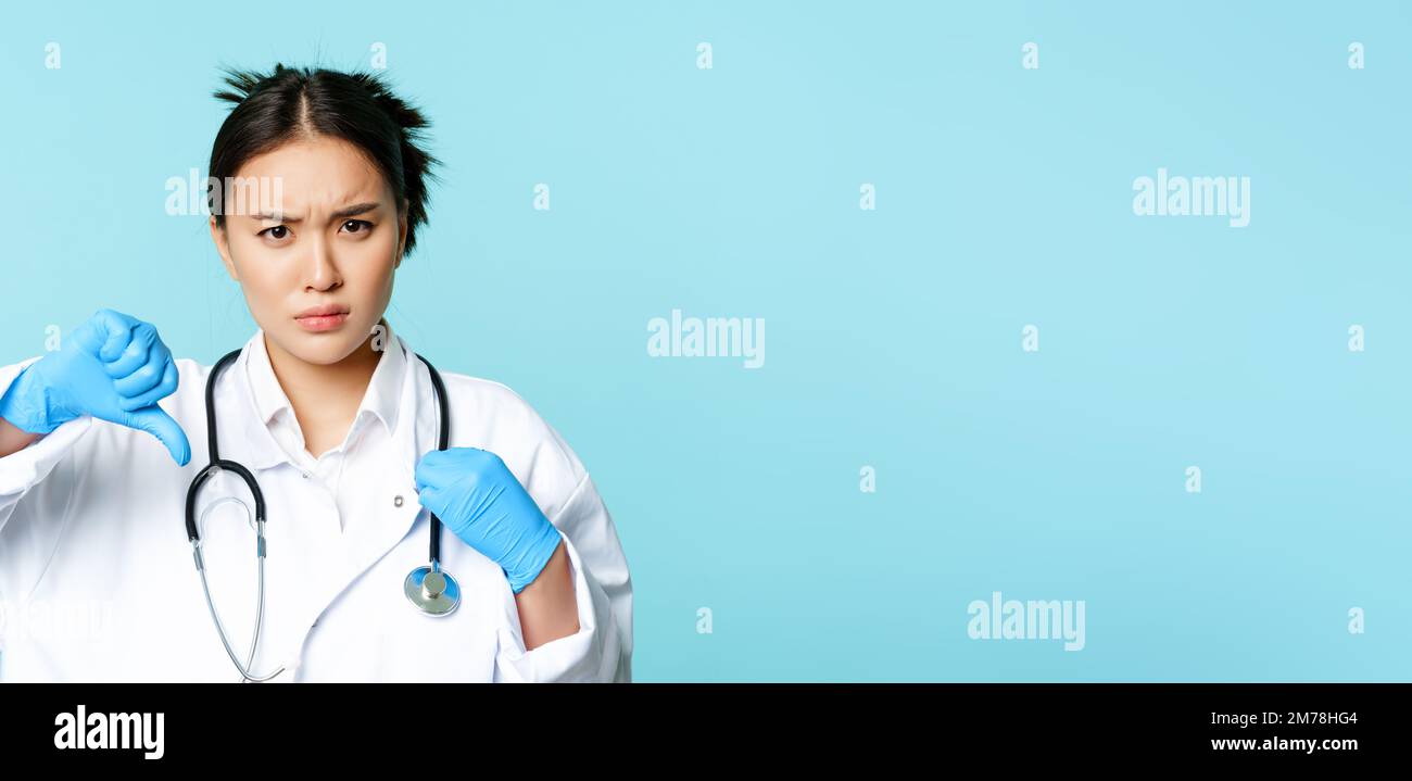 Angry asian woman doctor, physician in uniform and gloves shows thumbs ...