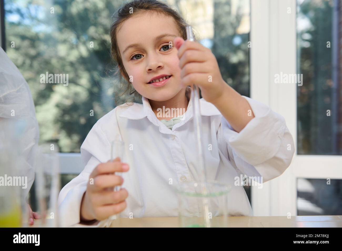 Close-up blue-eyed little girl in white lab coat, holds a graduated ...