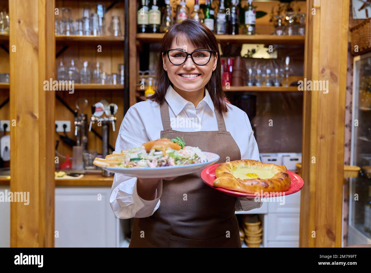 Middle aged female restaurant owner in apron with plates of cooked food ...