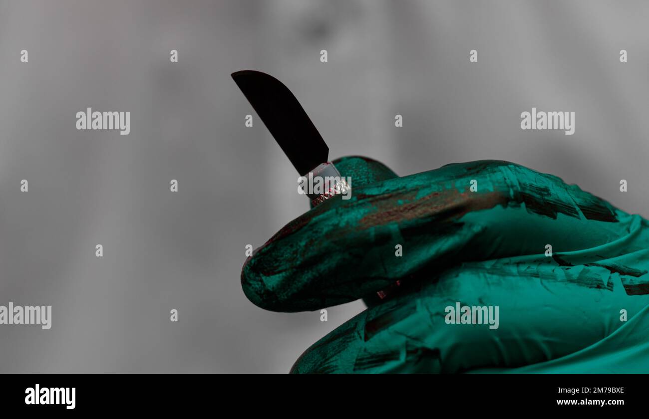 A hand in a green plastic glove holding a scalpel covered in blood ...