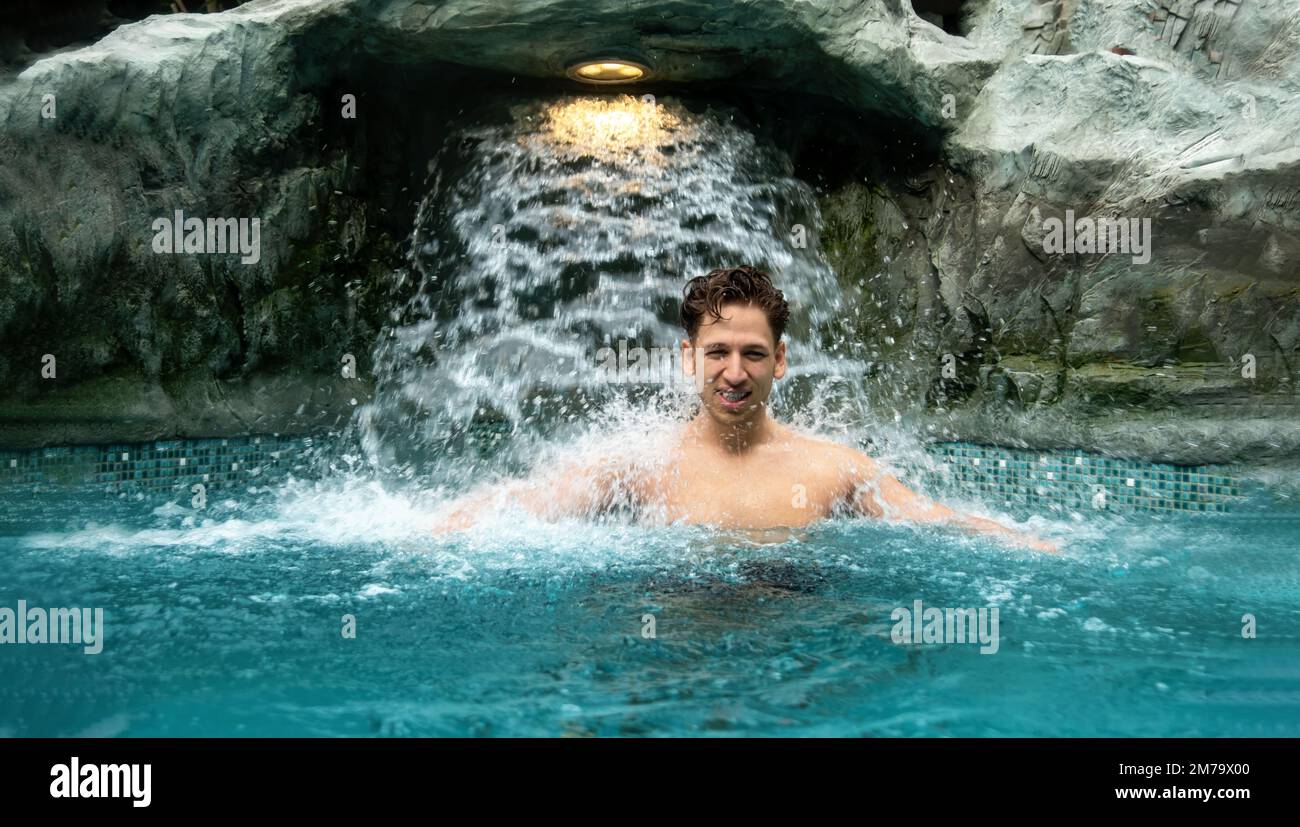 young man enjoys the splashing, falling water under the waterfall in ...