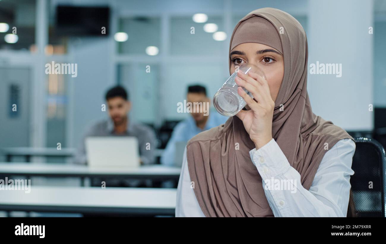 Thirsty businesswoman in hijab drinking pure mineral water in office, thoughtful dehydrated ...