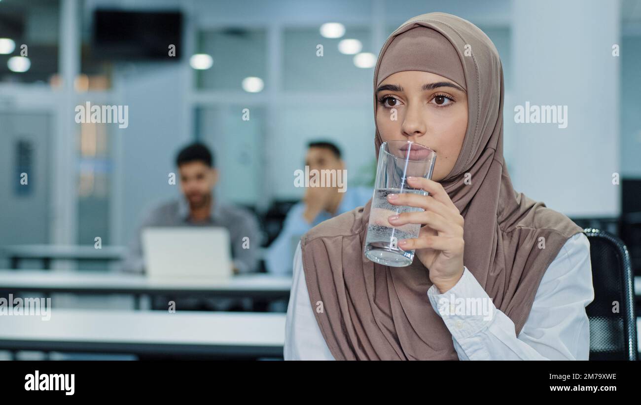 Thirsty millennial indian arabic female worker in hijab holding glass drinking pure mineral ...