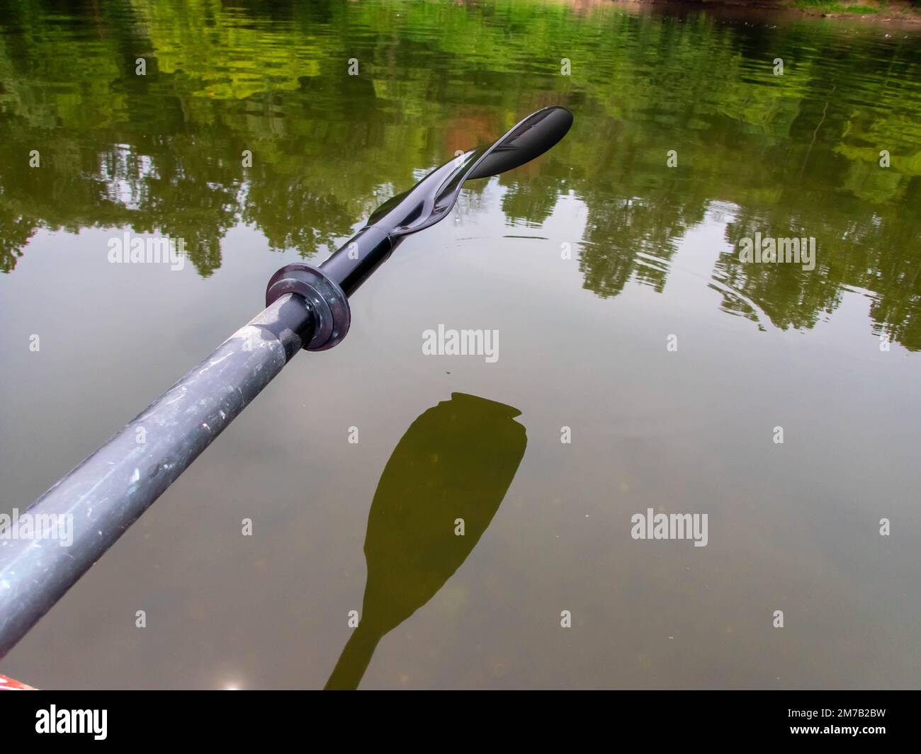 A raised kayak paddle above a woodland river with trees reflected in ...