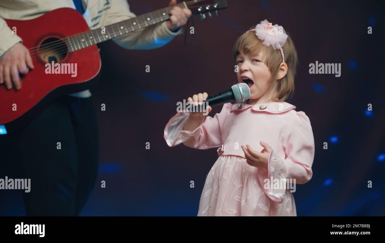 Llittle girl in vintage dress sings on stage, her father plays acoustic ...