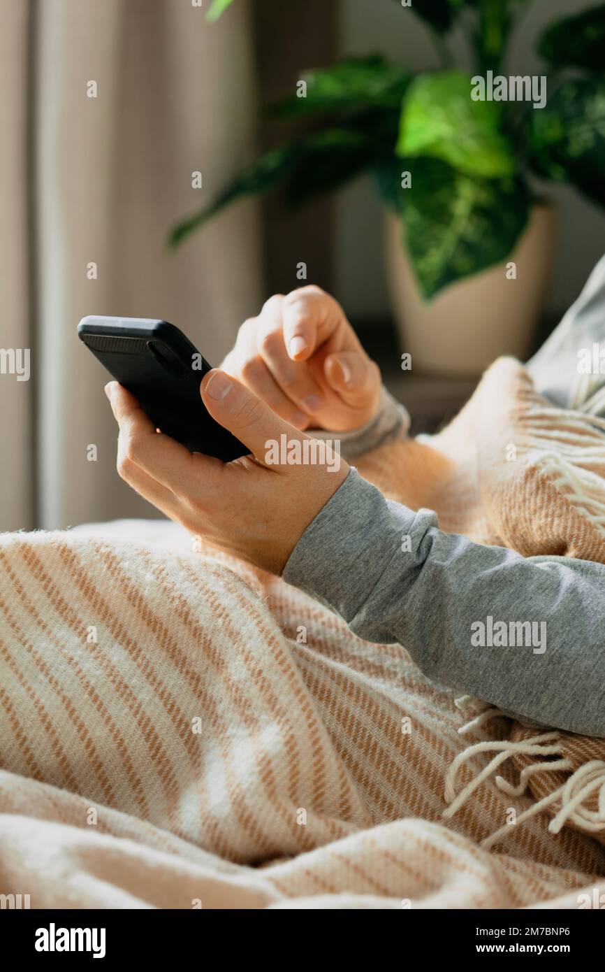 a caucasian man relaxing, using smart phone lying in bed under throw at ...