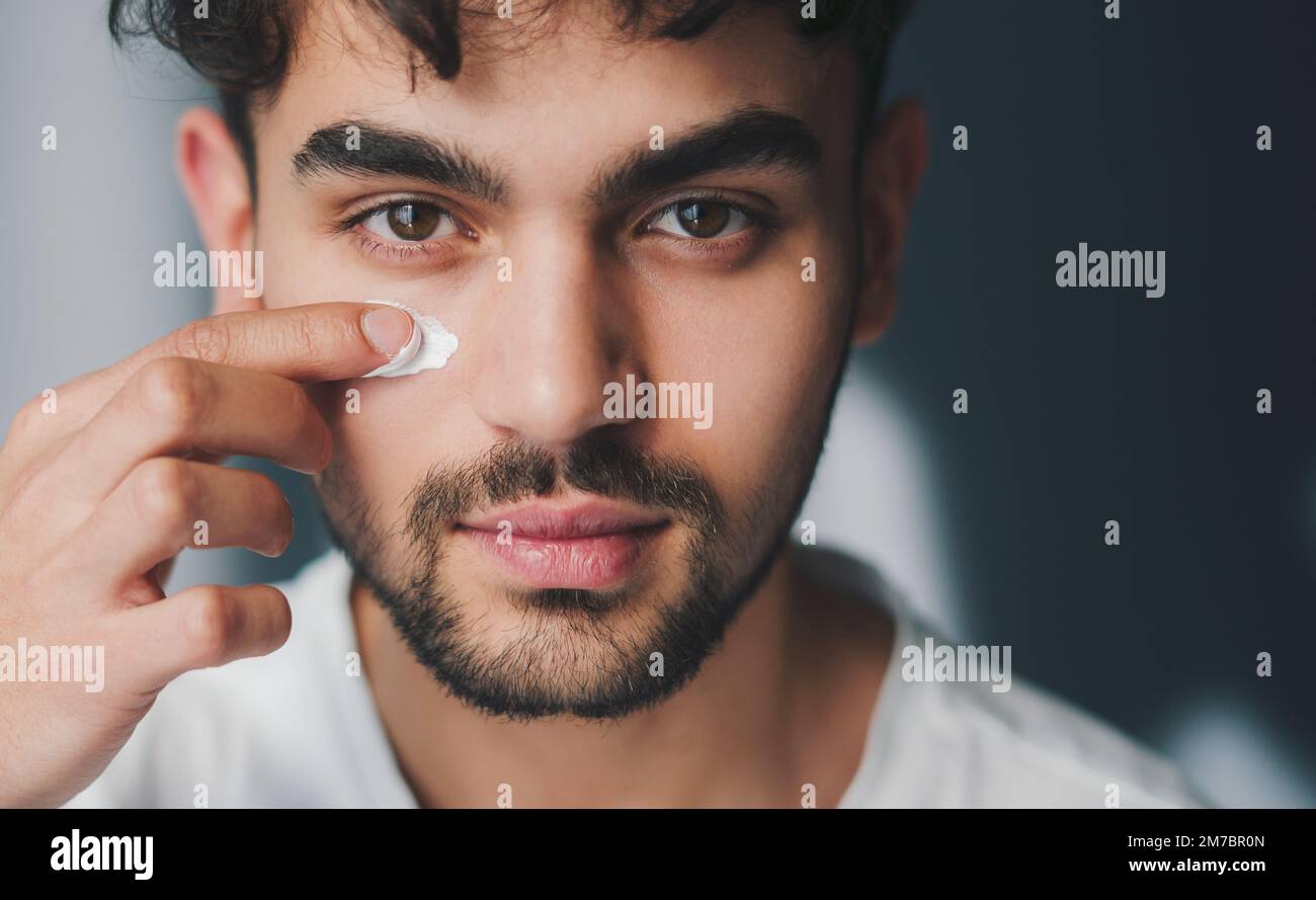 Close-up portrait of a young man sitting on sofa putting cream on face ...