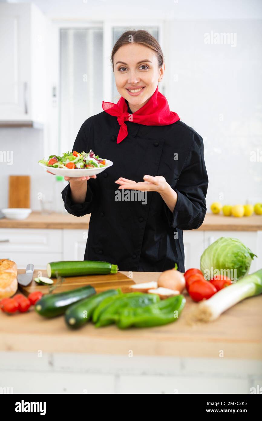 Smiling female chef in black uniform offering plate of vegetable salad ...