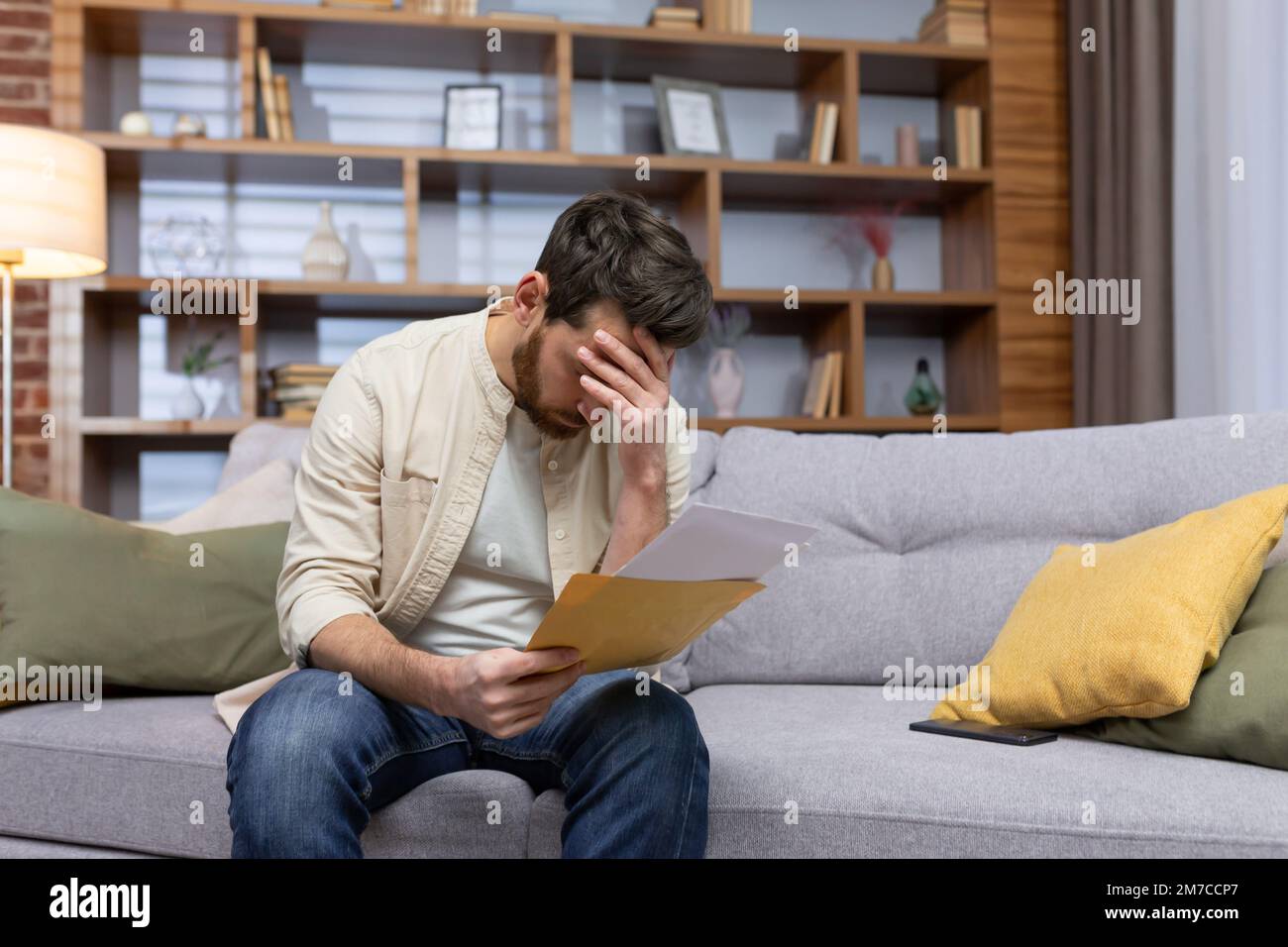 Upset man at home sitting on sofa reading letter with bad news ...