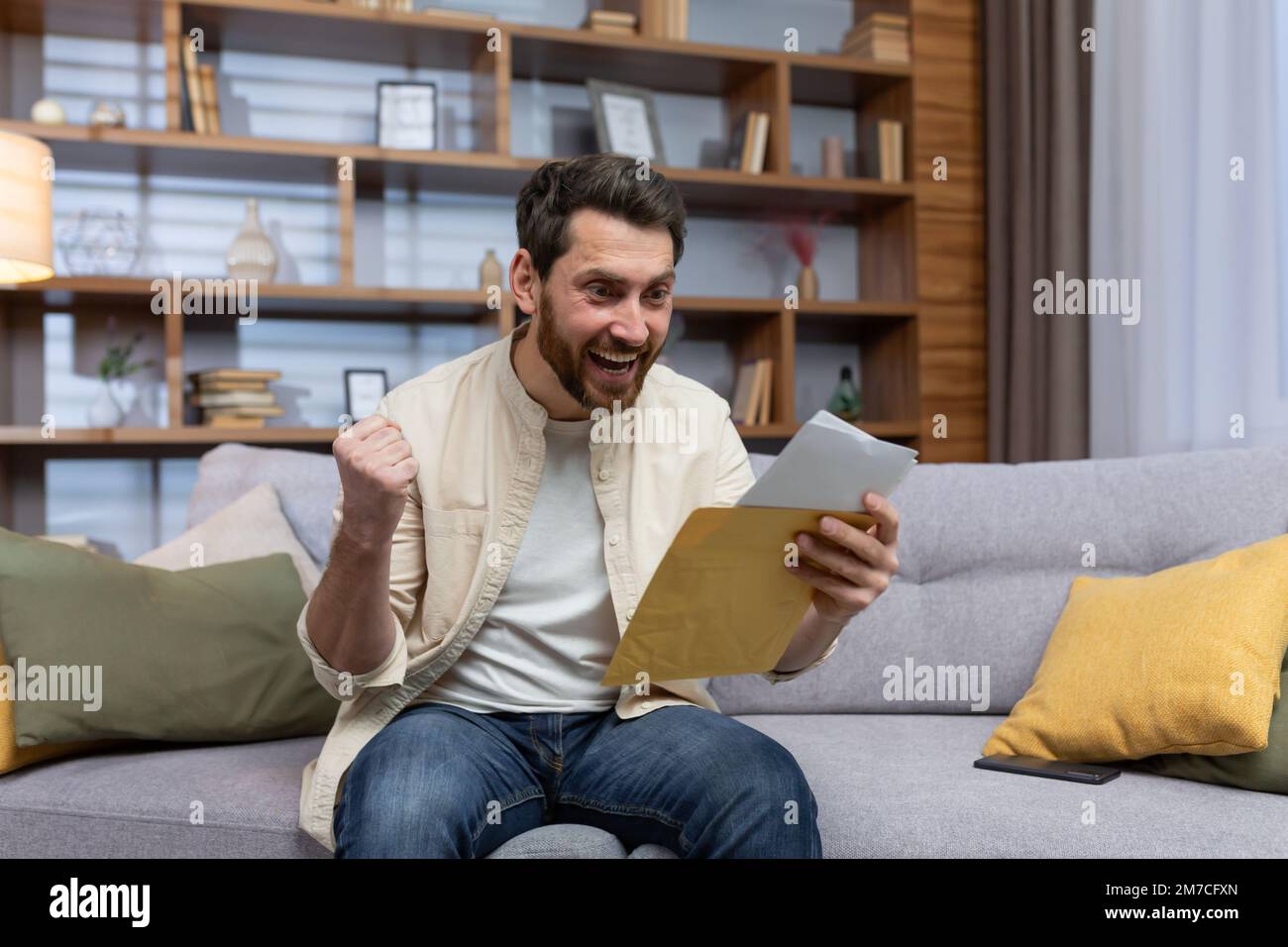 Joyful man at home holding envelope with notification message reading ...