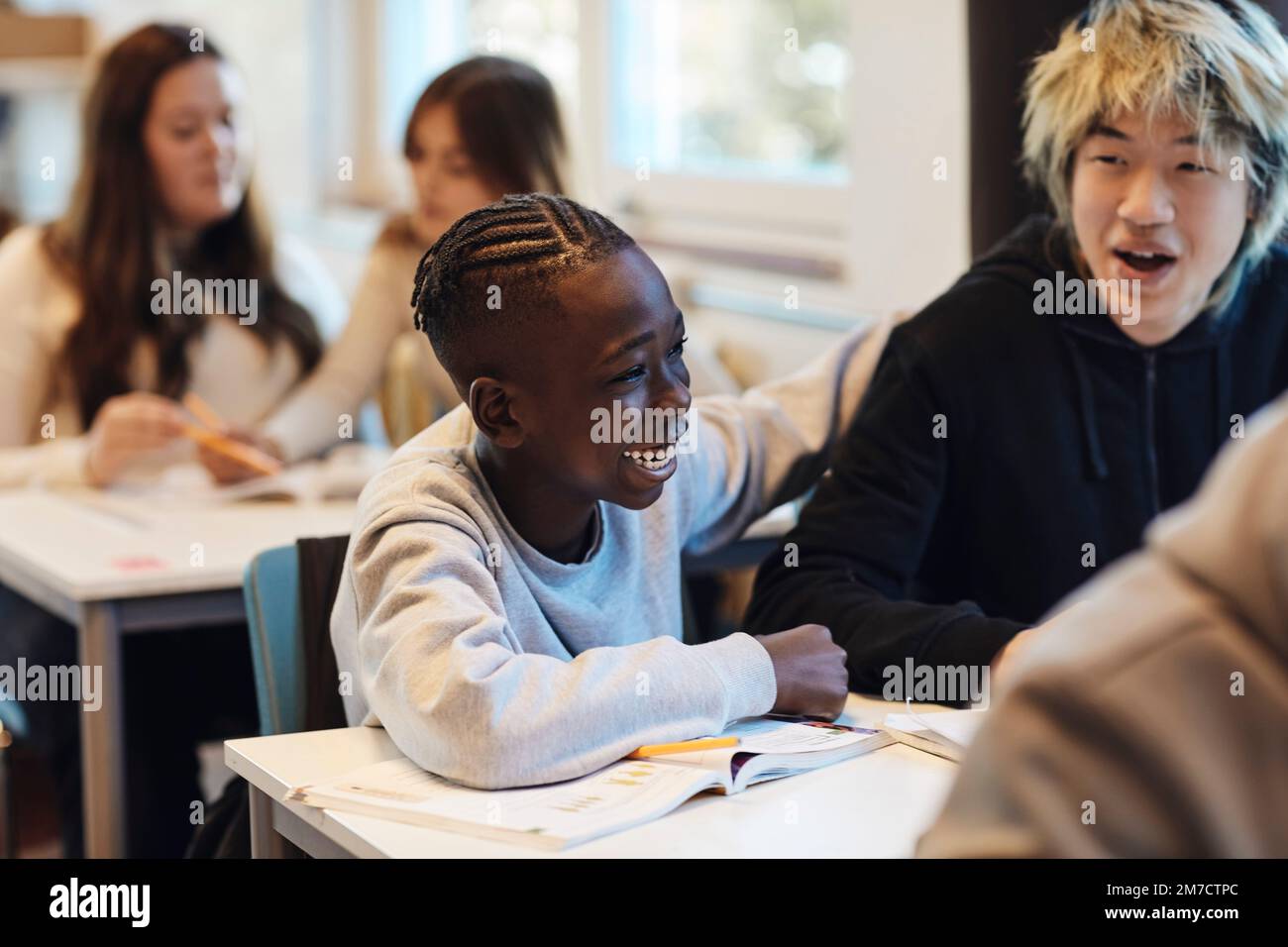 Happy teenage boy sitting by cheerful male friend at desk in classroom ...