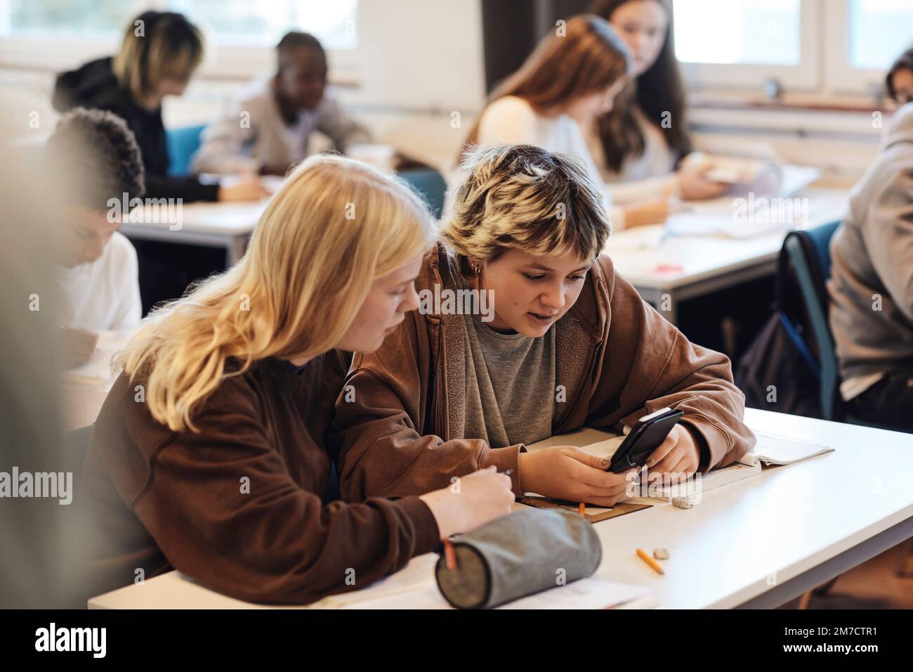 Teenage girl sharing smart phone with blond female friend sitting at ...