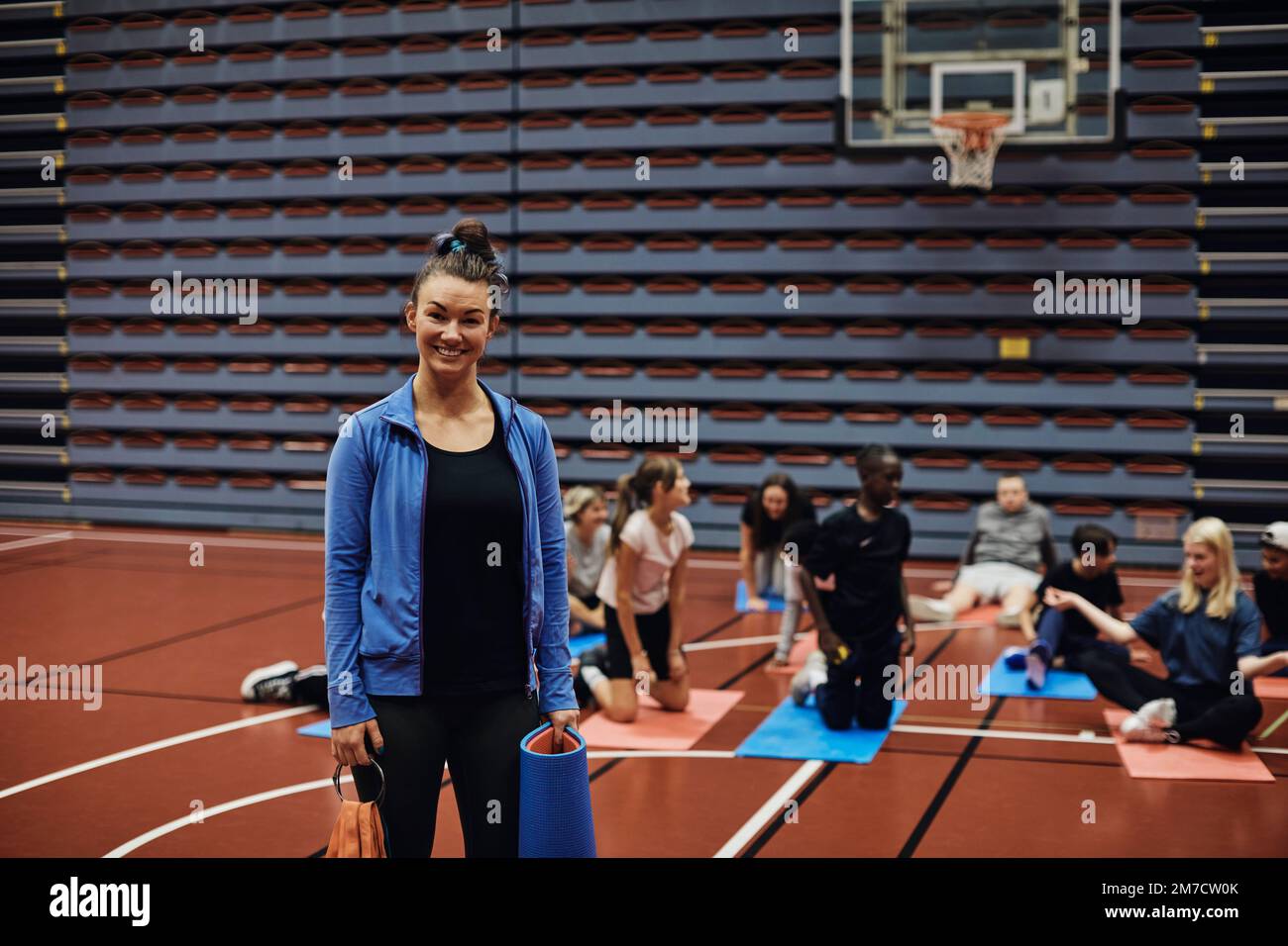 Portrait of smiling female coach standing with exercise mat against ...